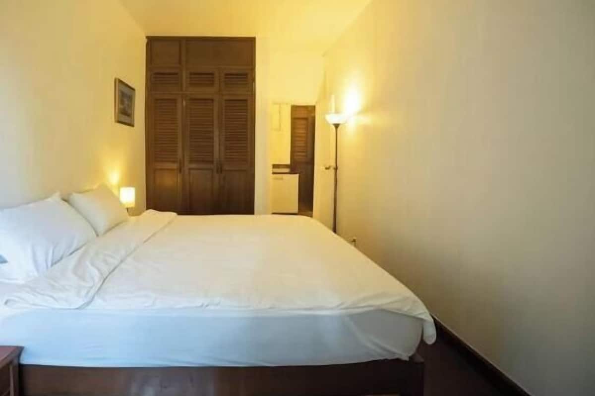 Uma cama de solteiro com cabeceira de madeira, uma mesinha de cabeceira e um criado-mudo. Há uma porta com painel de madeira e uma luminária fixada na parede.