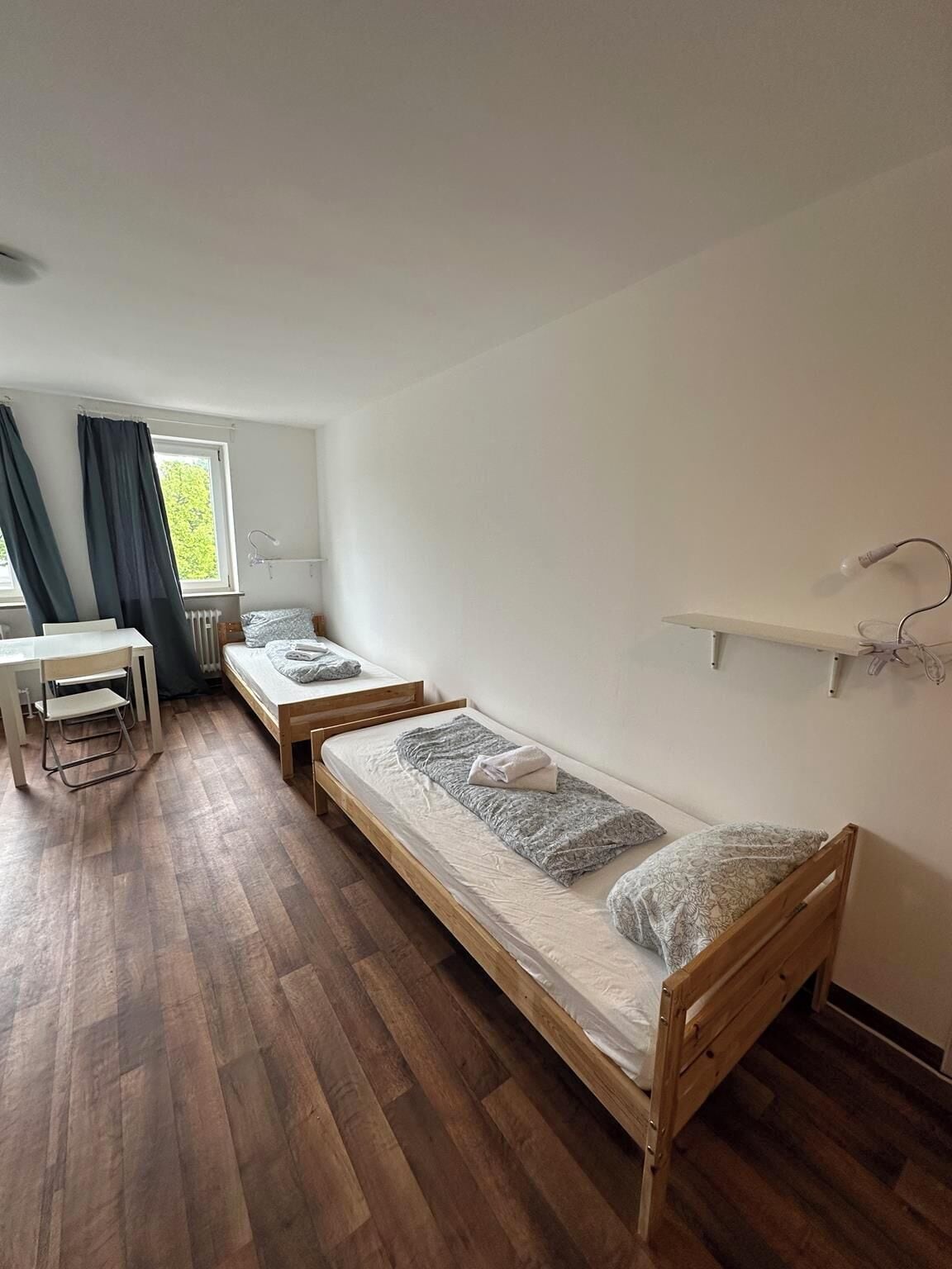 Quarto pequeno e arrumado, com duas camas individuais, piso de madeira, escrivaninha branca e janela com cortinas.