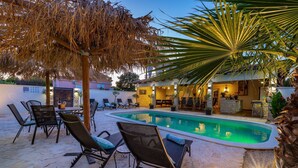 Uma área à beira da piscina com espreguiçadeiras, um pátio de pedra e um edifício com bar e piscina.
