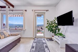Uma sala de estar moderna com um sofá, uma televisão de tela plana, uma janela grande com cortinas, um vaso de planta e vista para o horizonte da cidade.