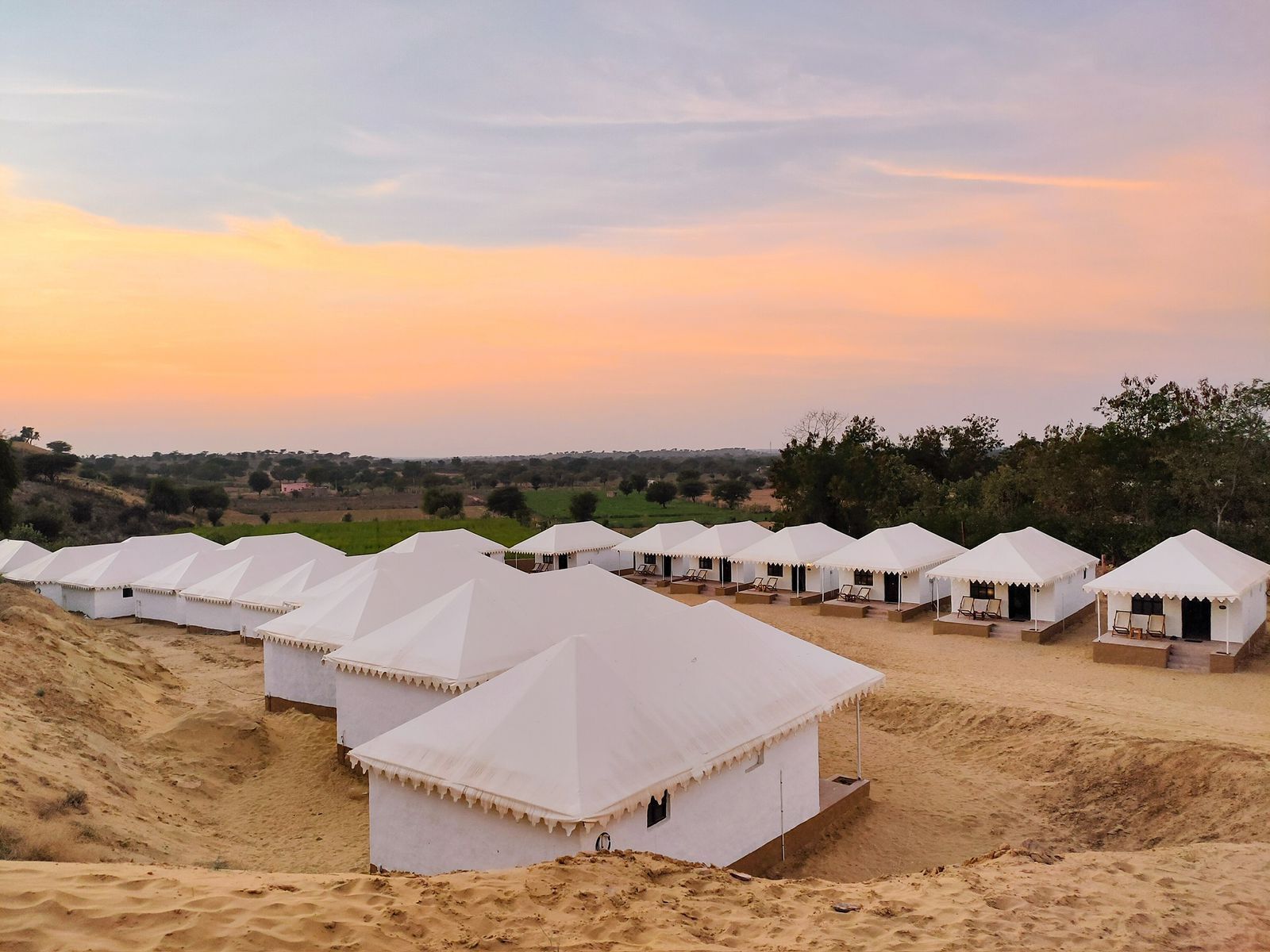 Fileira de tendas brancas com cadeiras de descanso, em paisagem desértica com um pôr do sol ao fundo.
