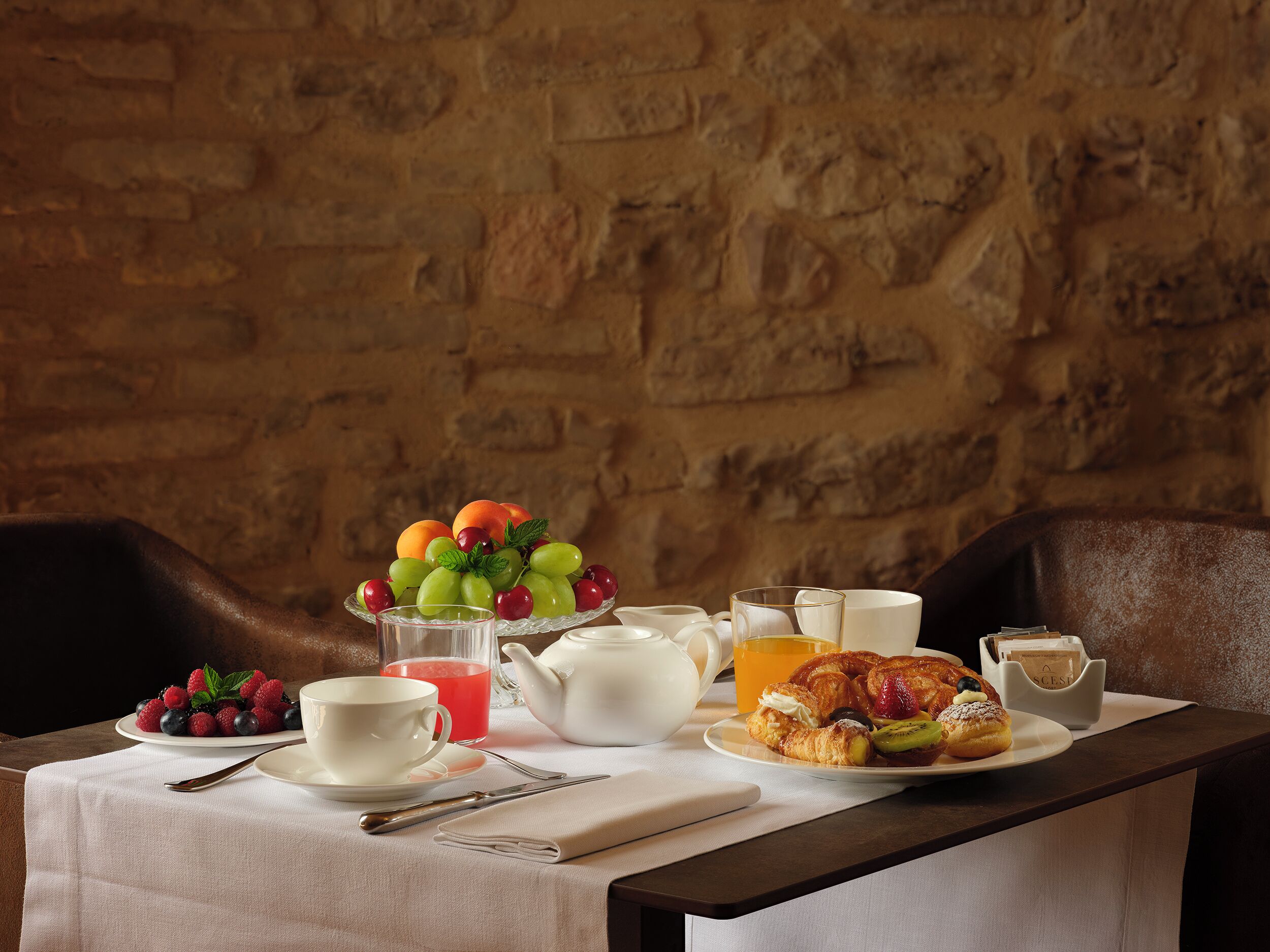 Mesa de café da manhã com arranjo de frutas, bule, xícaras e doces.