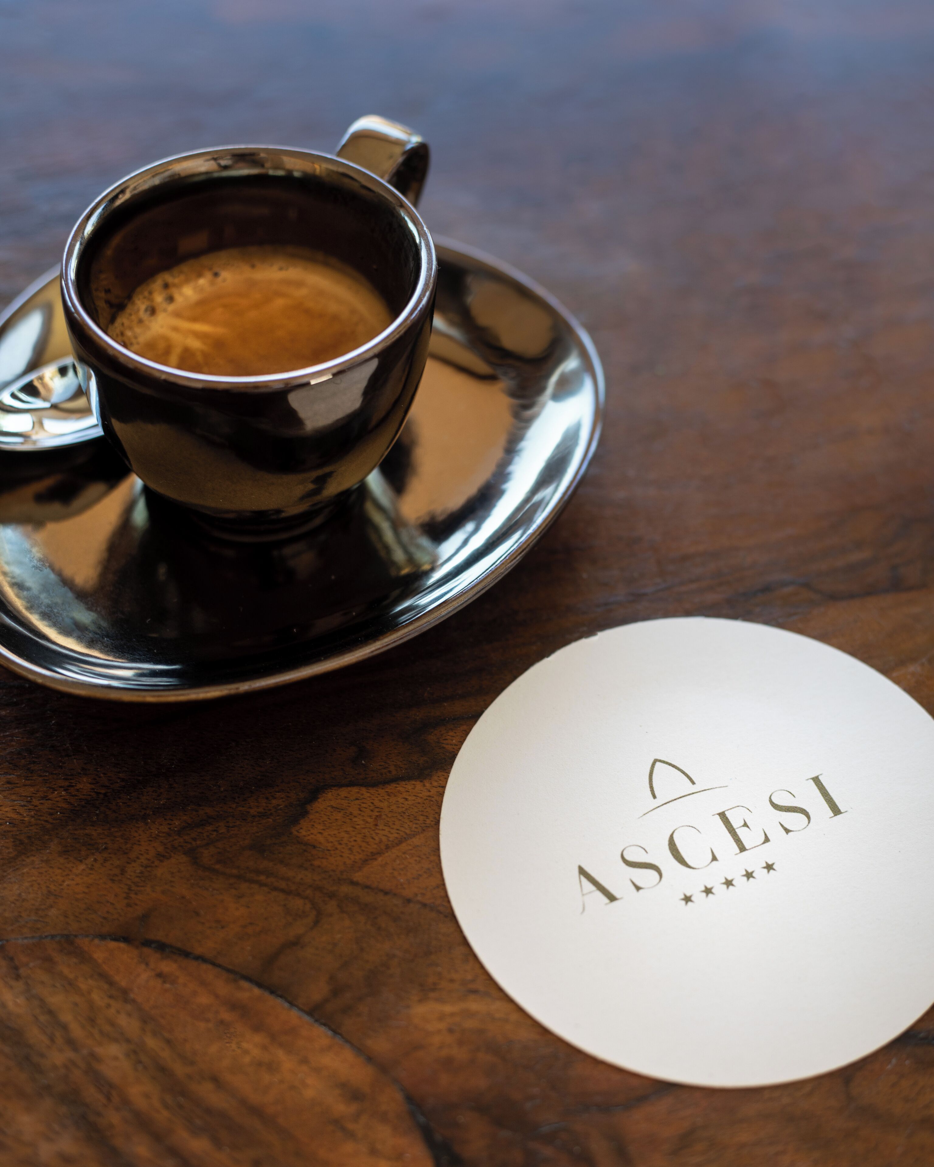 Uma chávena de café sobre um pires refletor, ao lado de um cartão de visita com o nome 'ASCESI' e quatro estrelas.