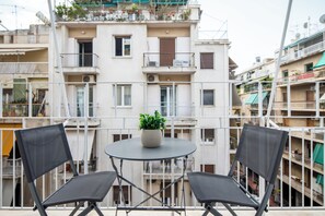 Uma varanda com duas cadeiras, uma mesa, um vaso de planta e um edifício com várias varandas ao fundo.