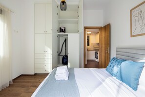 Quarto com cama, guarda-roupa, aspirador de pó e cozinha ao fundo.
