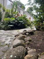 Uma trilha de pedra ladeada por pedras grandes e vegetação exuberante, conduzindo a um edifício com janelas amplas.