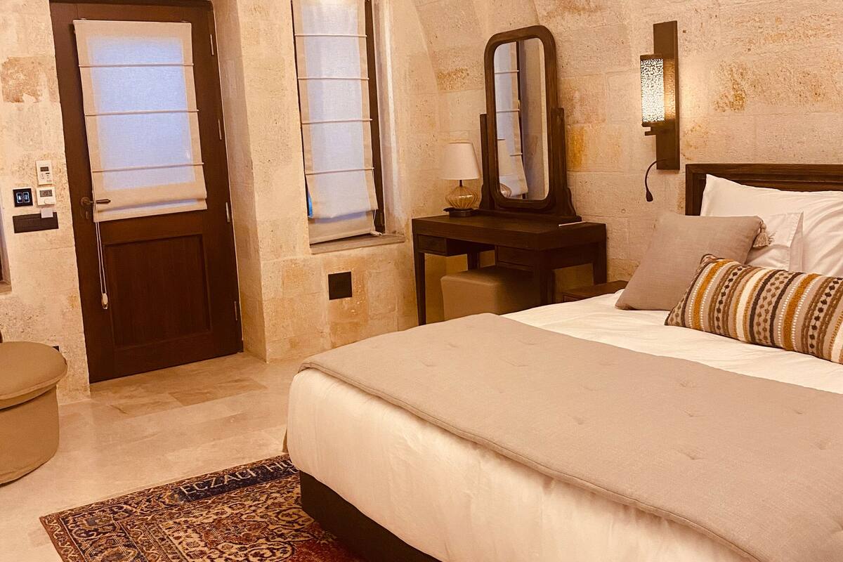 Quarto com cama, mesa de cabeceira, espelho, porta e janela com persianas.