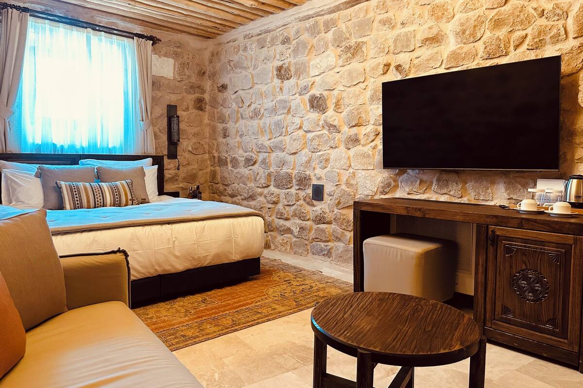 Quarto com parede de pedra, cama com várias almofadas, televisão, espaço para café e uma pequena mesa redonda.