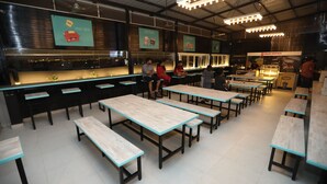 Restaurante com mesas longas comunitárias e mesas altas, com vista para a cidade através de grandes janelas.