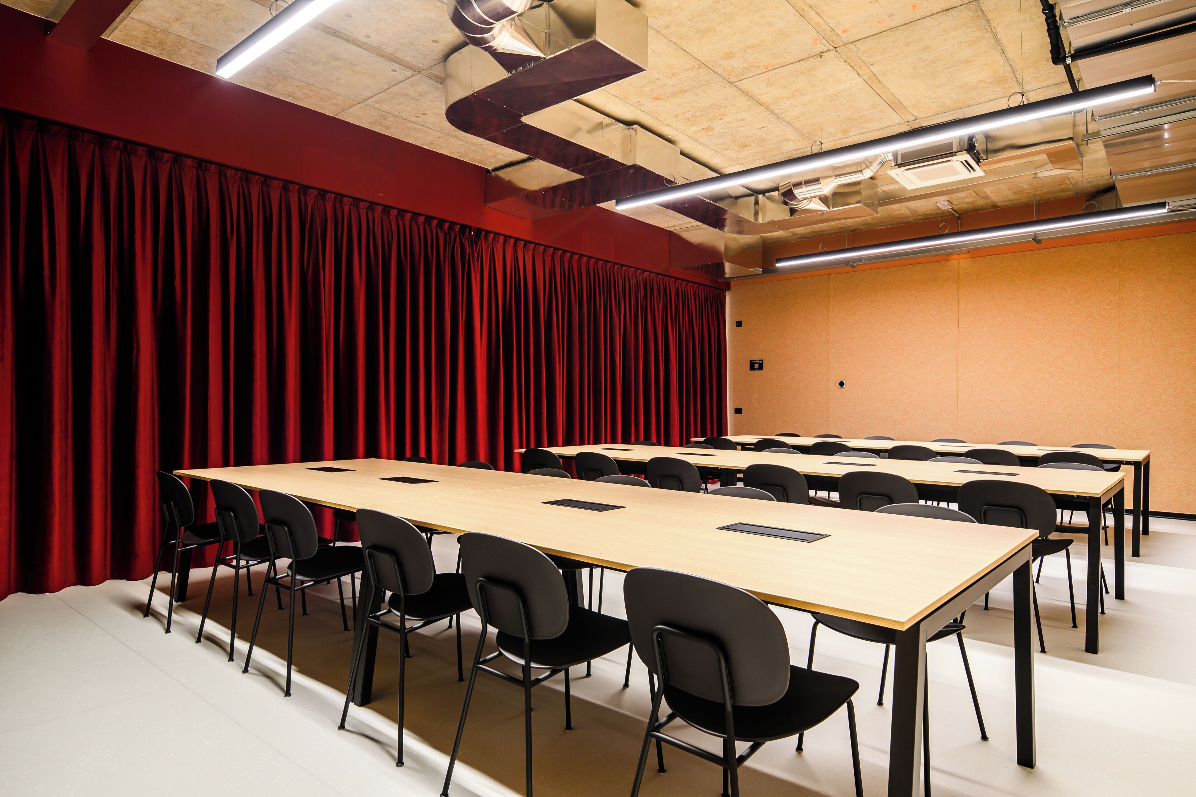 Uma sala de conferências com mesas longas e cadeiras, um pano de fundo com cortina vermelha e uma parede de madeira.