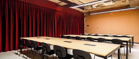 Uma sala de conferências com mesas longas e cadeiras, um pano de fundo com cortina vermelha e uma parede de madeira.