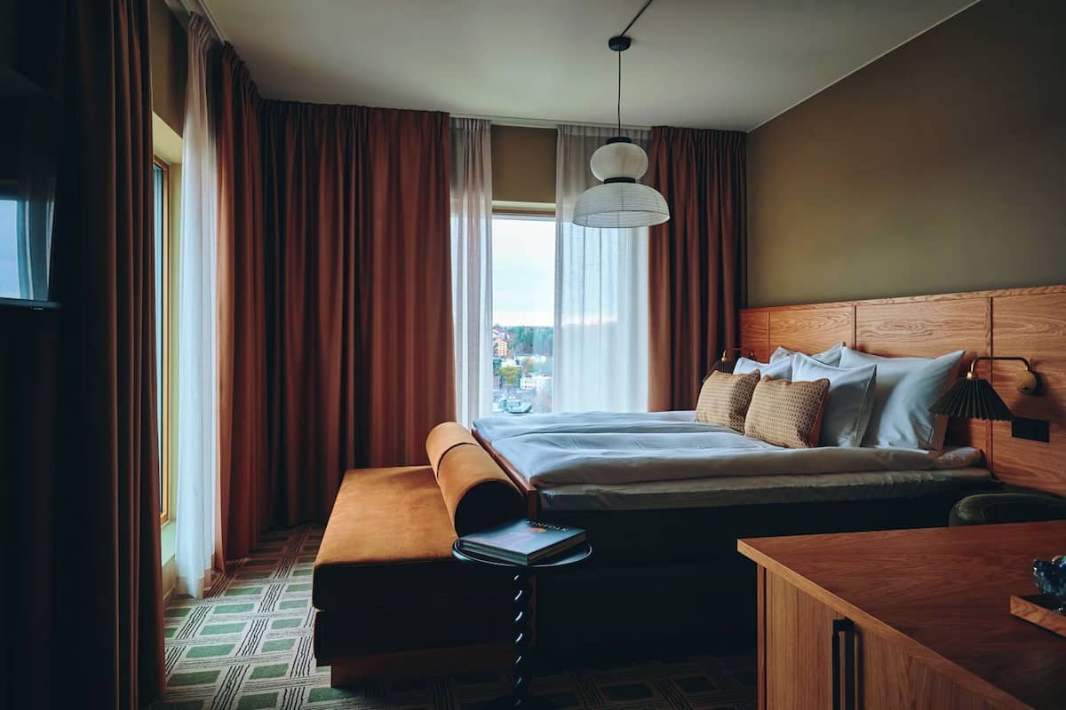 Quarto de hotel com uma cama grande, cabeceira de madeira, escrivaninha e vista para a cidade pela janela.