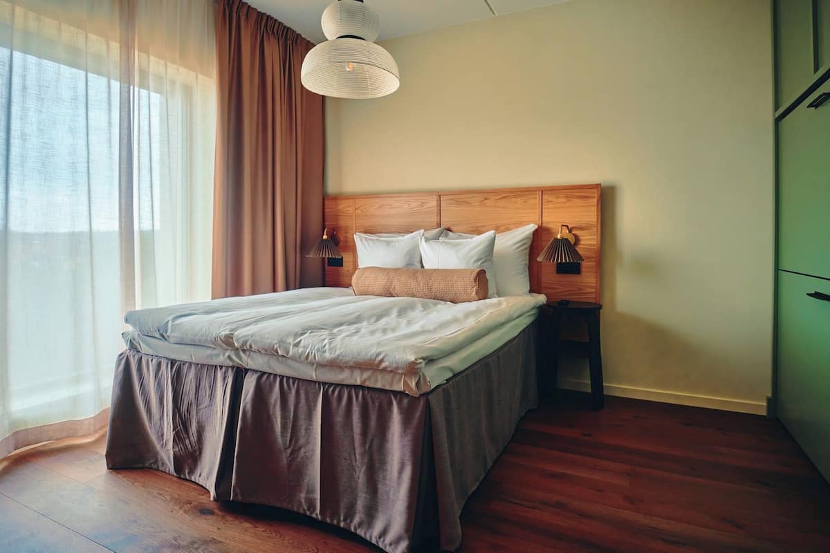 Quarto bem organizado com cabeceira de madeira, cama com colcha cinza, mesa de cabeceira com abajur e guarda-roupa verde.