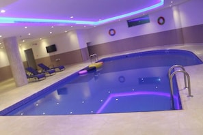 Uma piscina interior com tonalidade azul, rodeada por espreguiçadeiras e um design de teto moderno.