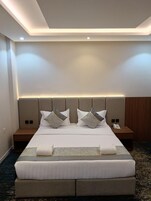 Quarto de hotel com cama grande, iluminação na cabeceira, duas mesas de cabeceira e piso coberto por carpete.