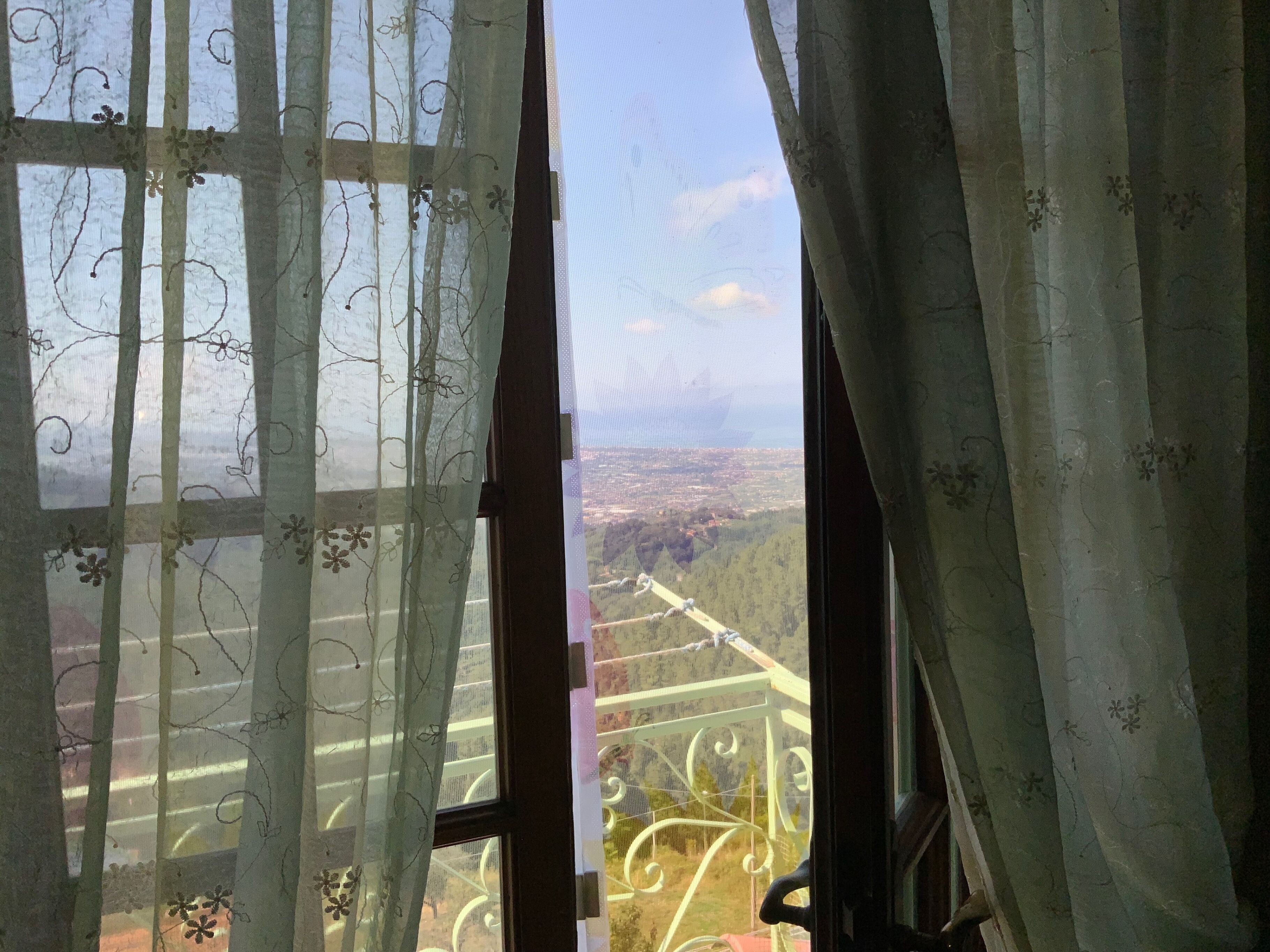 Vista pela janela com cortinas florais, mostrando uma varanda com grade de metal e paisagem distante.