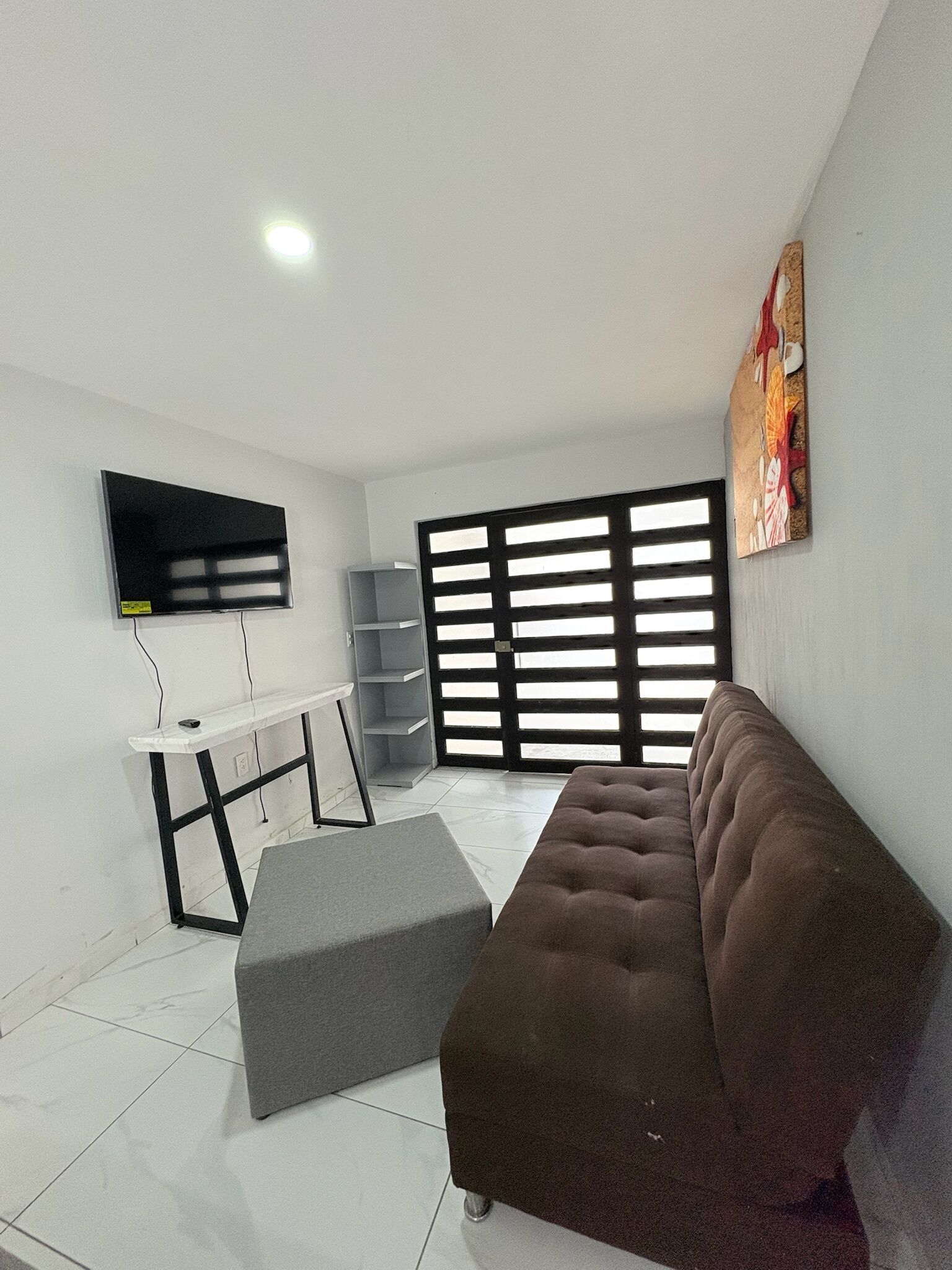 Uma sala de estar com um sofá marrom, um banquinho cinza, uma TV na parede, uma escrivaninha com um computador e uma janela com persianas.