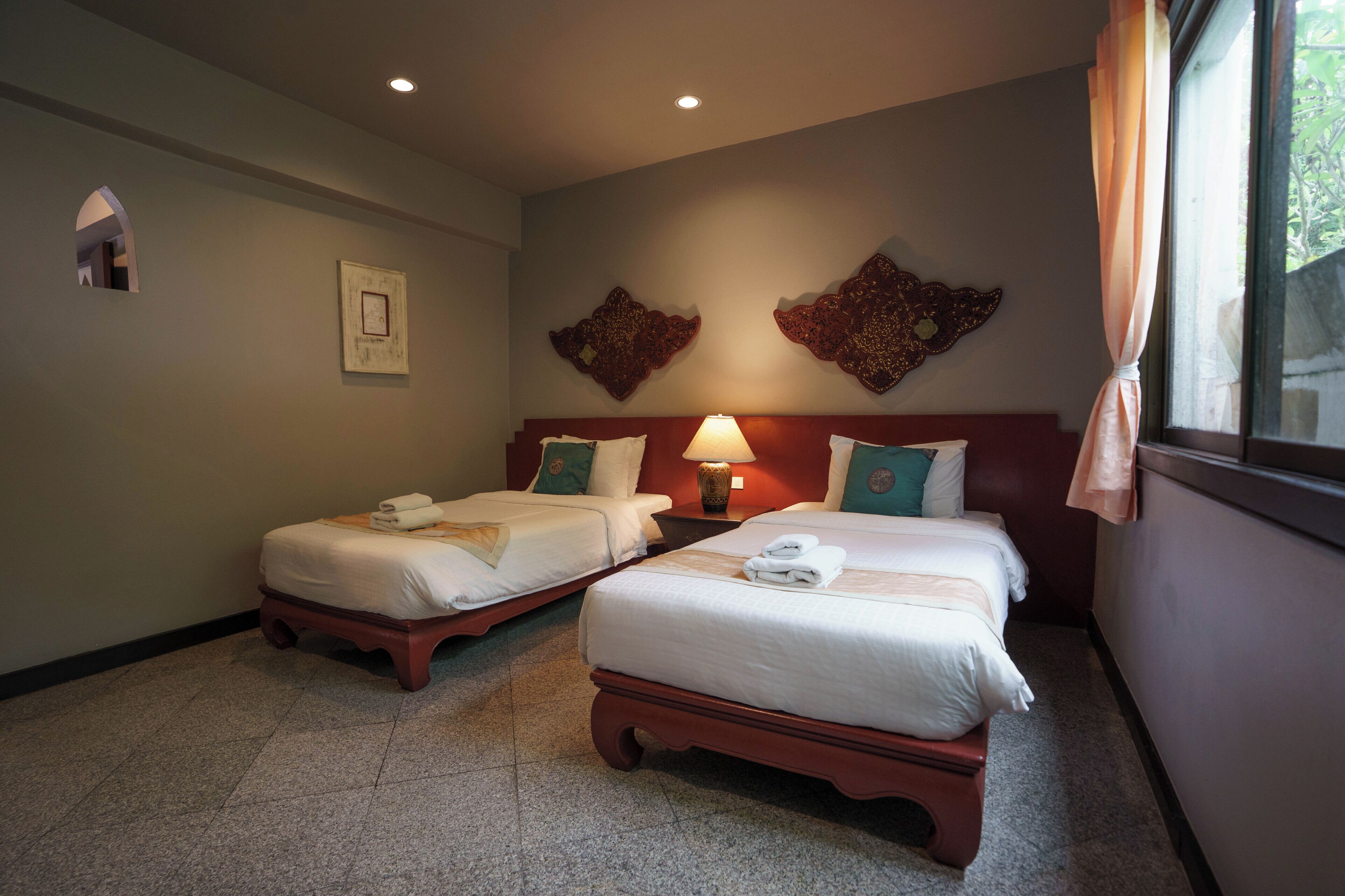 Quarto de hotel com duas camas, uma mesa de cabeceira com luminária, um quadro na parede e uma janela com cortinas.