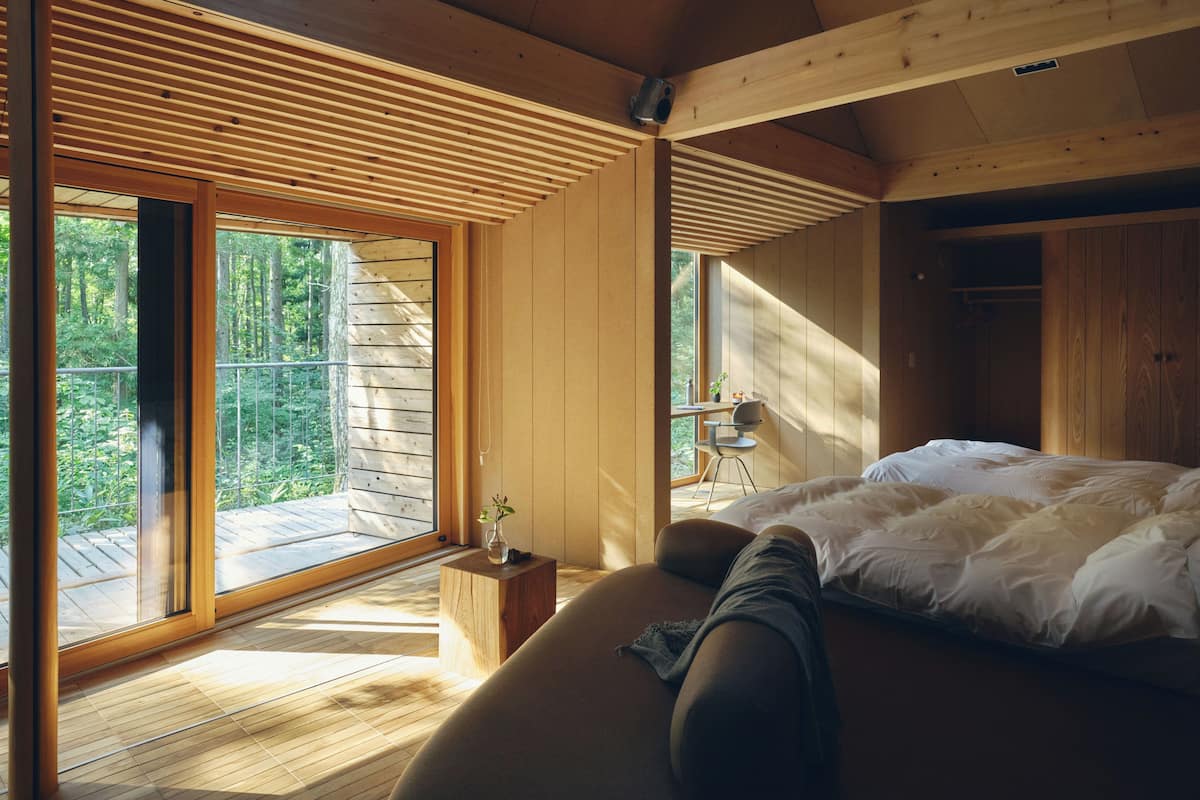 Quarto com cama grande, piso de madeira, escrivaninha com cadeira e janela com vista para uma floresta.