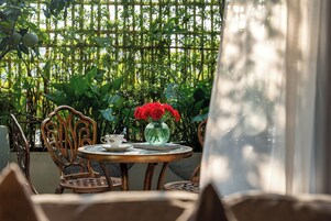 Uma área externa com espaço para sentar, incluindo uma mesa redonda, cadeiras, um vaso com flores vermelhas e um pano de fundo de folhagem verde.