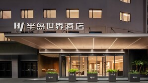 Entrada de hotel com design moderno, apresentando acesso coberto, portas de vidro e plantas em vasos.