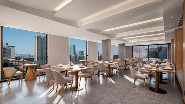 Uma sala de jantar moderna com mesas, cadeiras, janelas amplas e vista para a cidade.