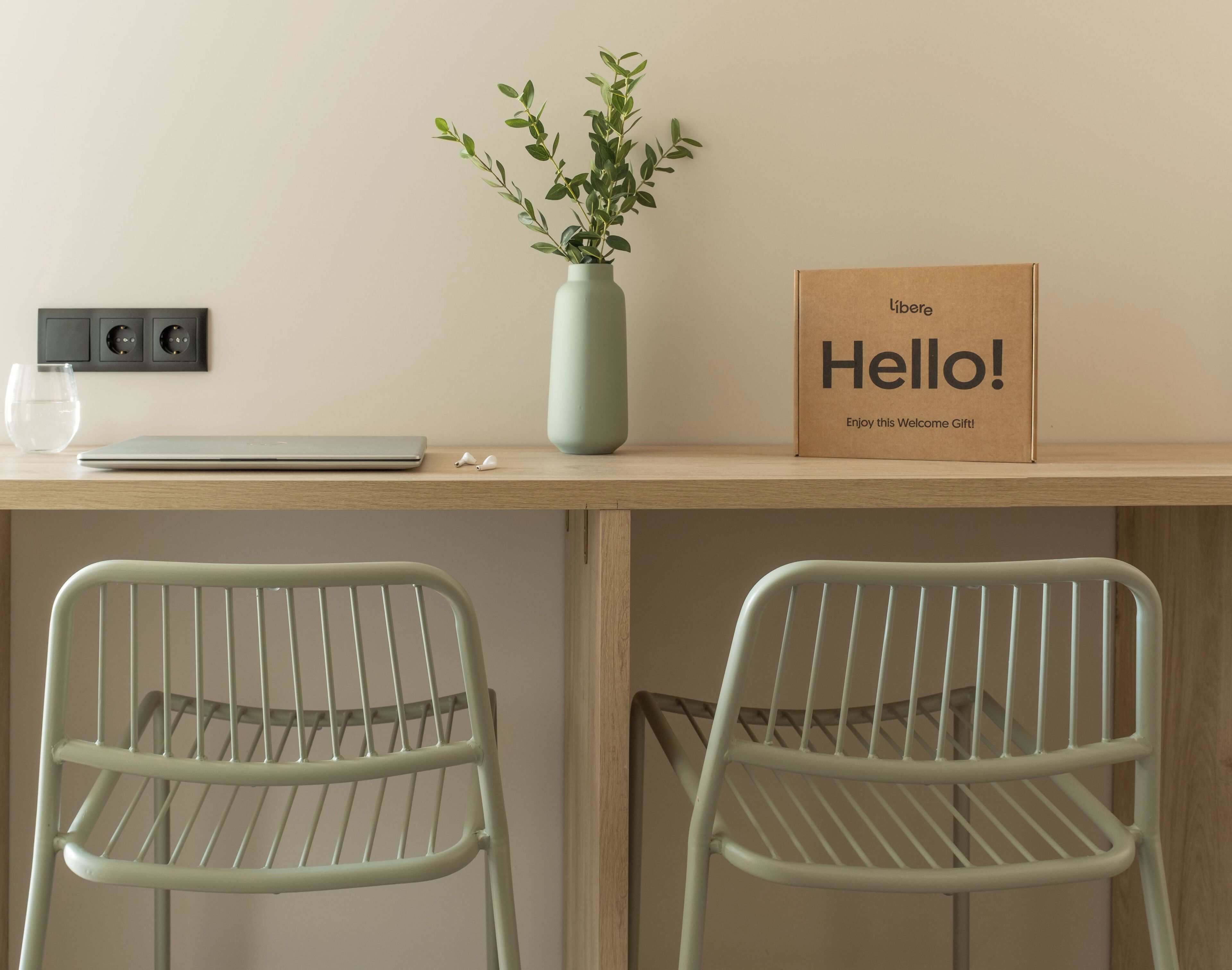 Uma mesa com um laptop, um vaso com folhas verdes, um aviso com a palavra 'Hello!' e duas cadeiras brancas.