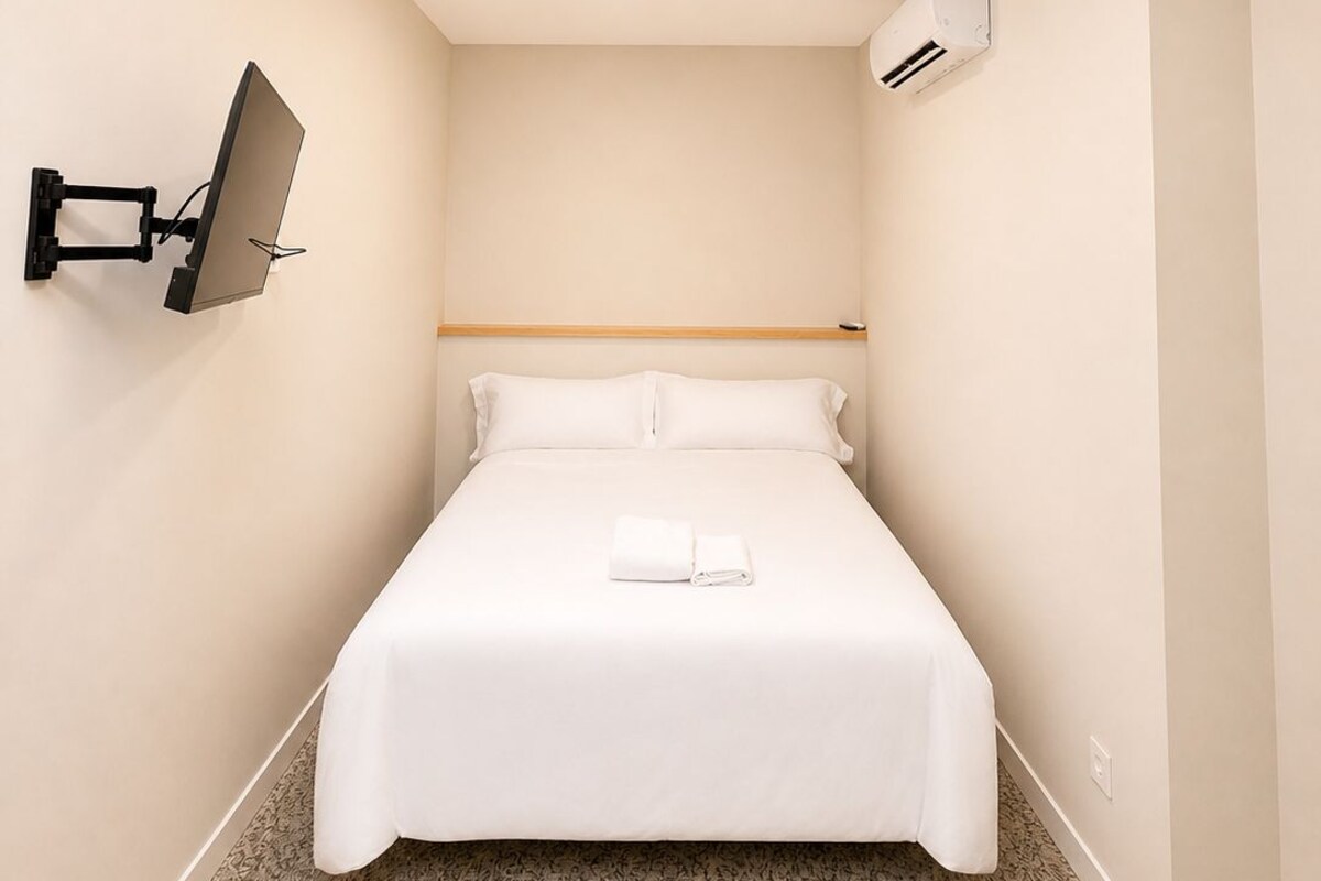 Quarto de hotel com cama, televisão na parede, aparelho de ar condicionado e claraboia.