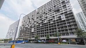 Um edifício alto com vários andares e varandas, uma rua com carros e um ponto de ônibus.