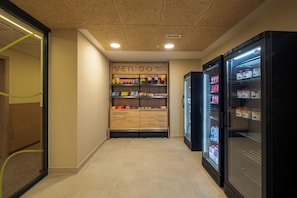 Um corredor com uma máquina de vendas automática, um balcão refrigerado e uma prateleira de madeira com lanches.