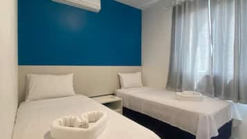 Quarto de hotel com duas camas, parede azul como detalhe, aparelho de ar condicionado, mesa de cabeceira e janela com cortinas leves.