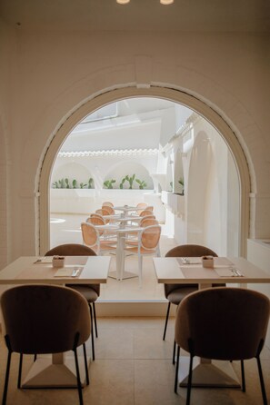 Uma área de refeições com mesas e cadeiras brancas, um arco de entrada que leva a um pátio externo com plantas.