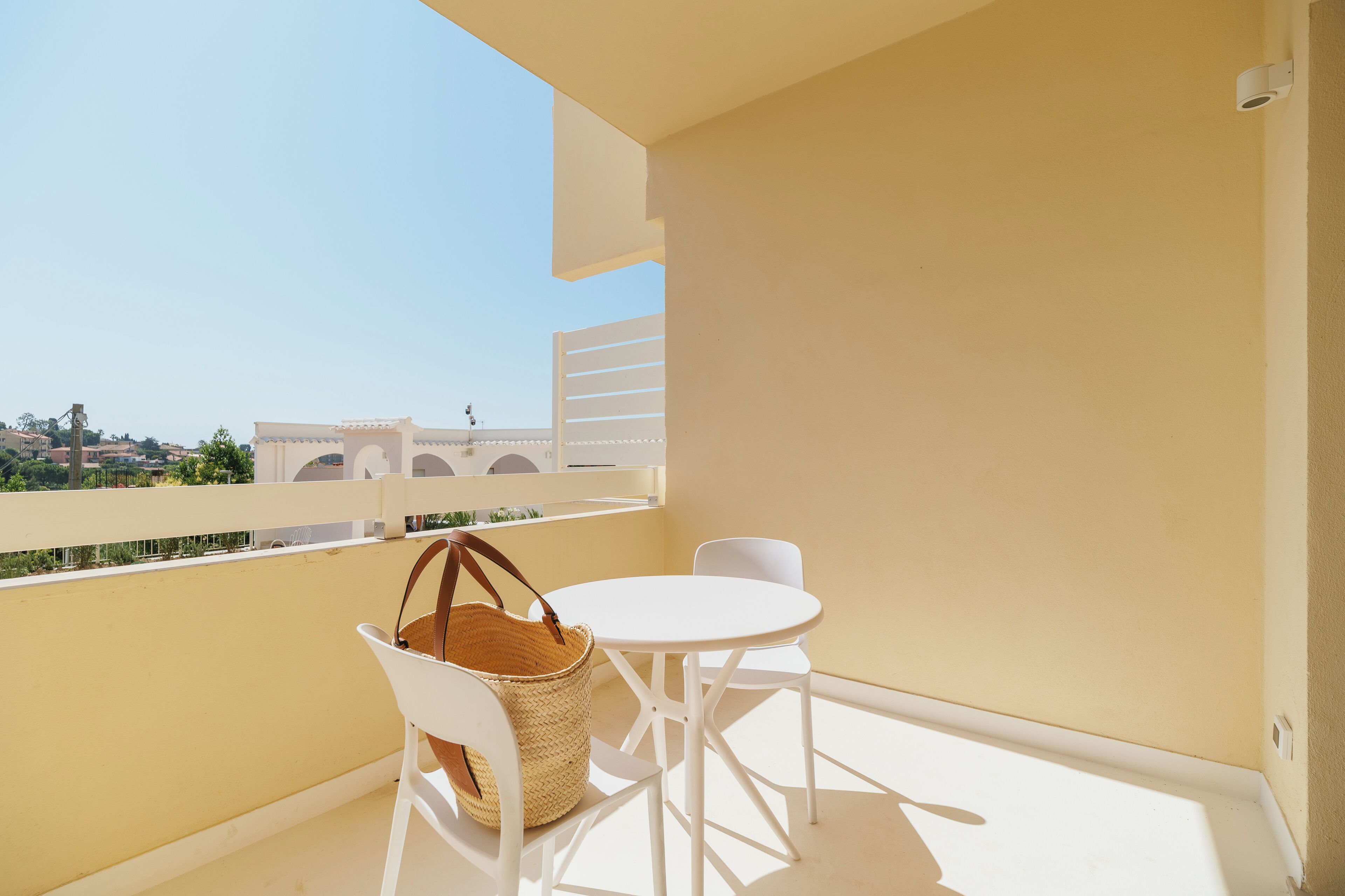 Varanda com mesa e cadeiras brancas, uma cesta de vime, e vista para uma ponte e edifícios ao fundo.