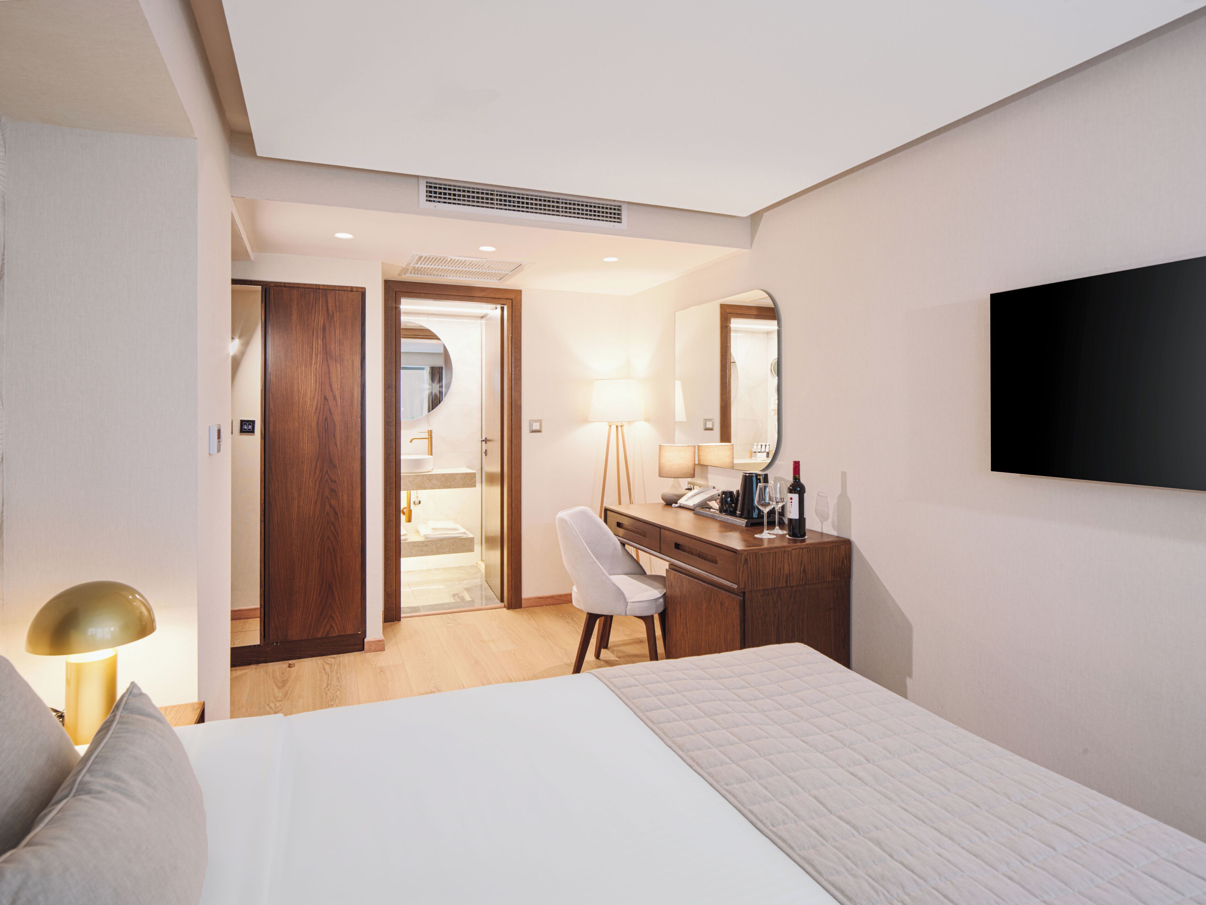 Quarto de hotel com cama, escrivaninha com espelho, televisão na parede e banheiro visível através de uma porta aberta.