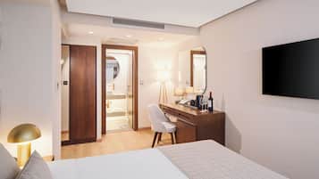 Quarto de hotel com cama, escrivaninha com espelho, televisão na parede e banheiro visível através de uma porta aberta.