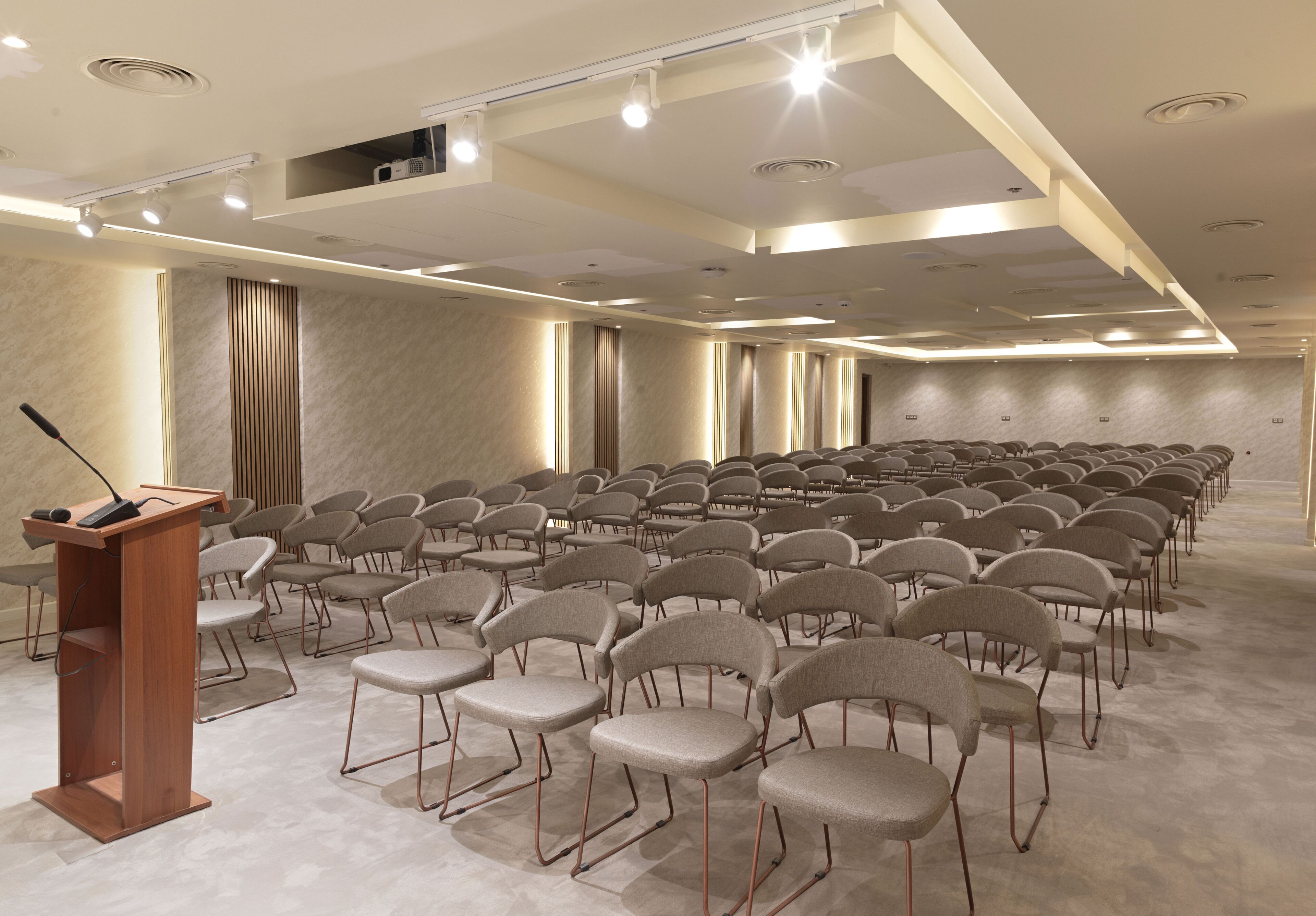 Sala de conferências com fileiras de cadeiras, um pódio e iluminação embutida no teto.