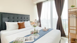 Uma cama bem arrumada com cabeceira estofada, uma mesa de cabeceira com um vaso, e vista para a cidade através de grandes janelas com cortinas escuras.