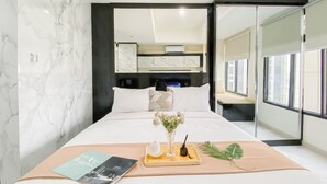 Quarto de hotel moderno com parede de mármore, cama com roupa de cama branca, mesa de cabeceira com um livro e um vaso de flores, e uma grande janela com vista.