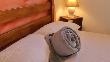 Uma cama bem arrumada com uma toalha enrolada, cabeceira de madeira, mesa de cabeceira com abajur e parede com cortina.