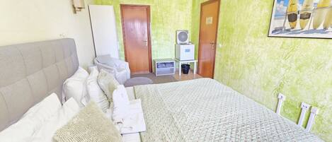 Quarto de hotel com cama, poltrona, escrivaninha, ventoinha e porta.