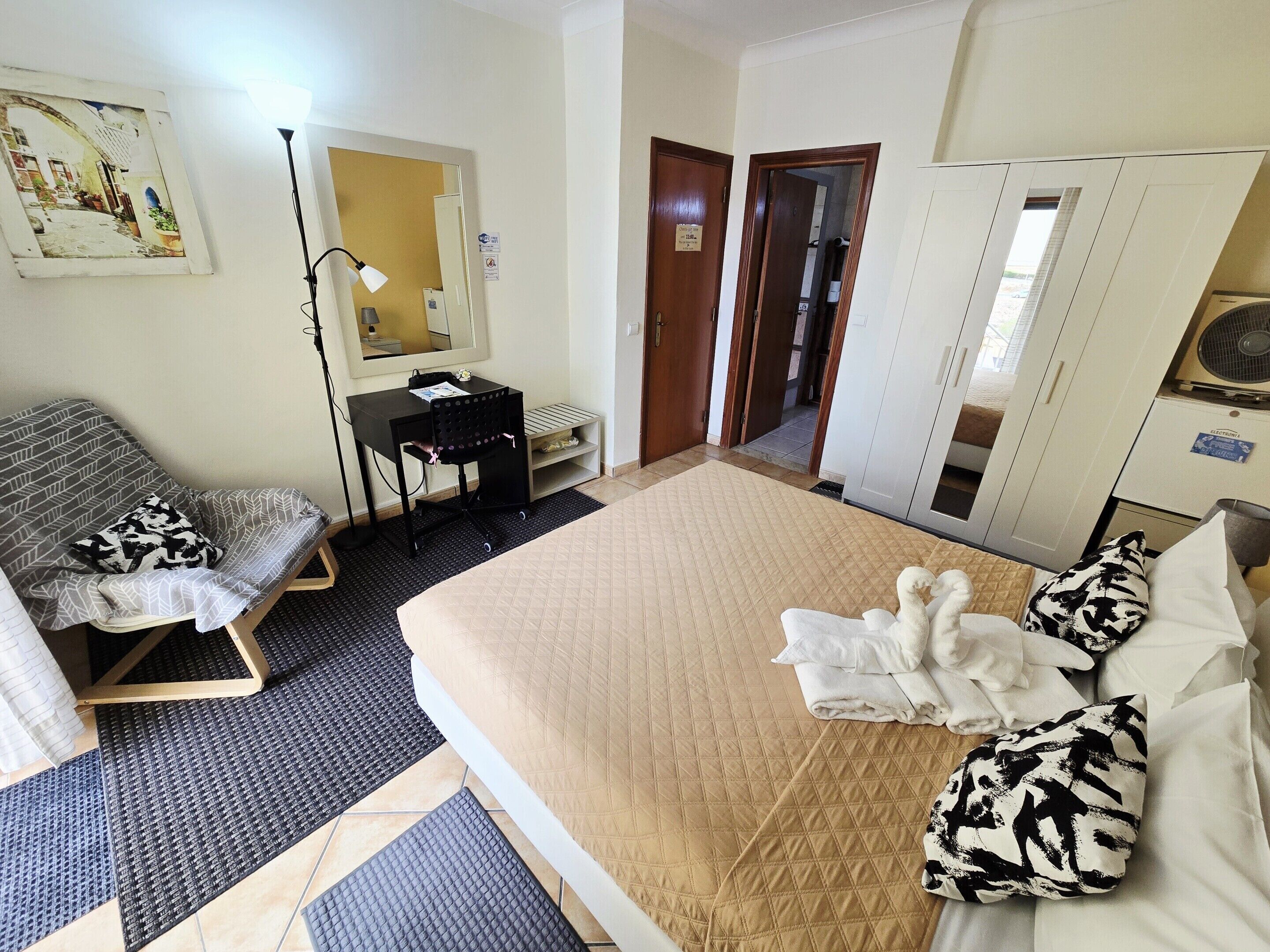 Quarto de hotel com uma cama, escrivaninha, cadeira, guarda-roupa e espelho.