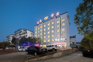 Um hotel com vários andares, uma fileira de janelas na fachada, um estacionamento com vários carros e uma entrada bem iluminada.