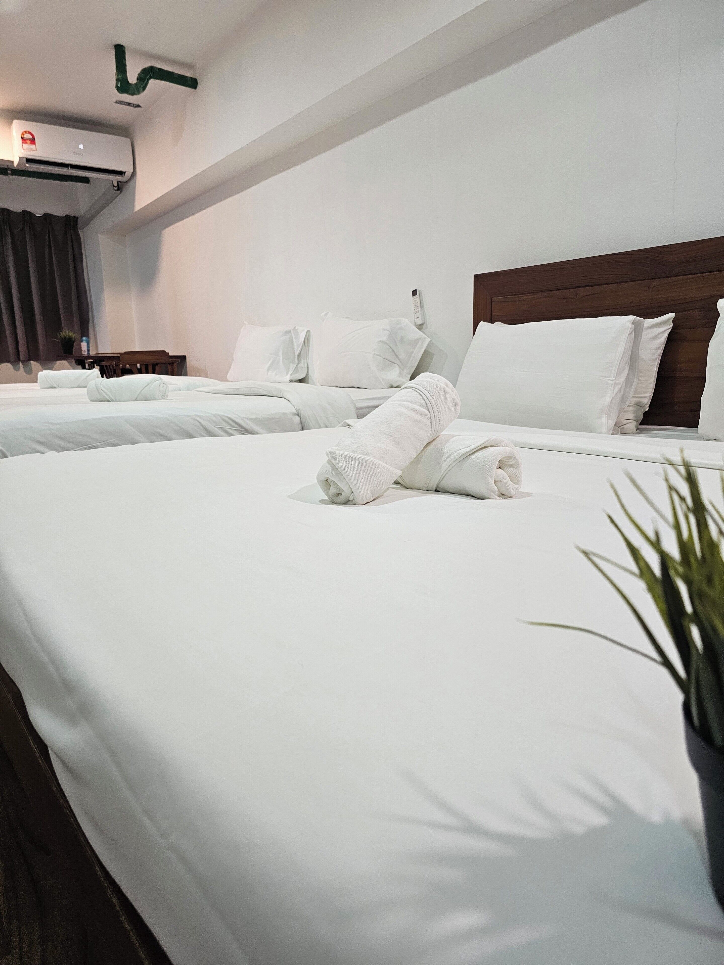 Quarto de hotel com duas camas, roupa de cama branca, cabeceira de madeira, mesa de cabeceira, ar condicionado fixado na parede e um vaso com planta.