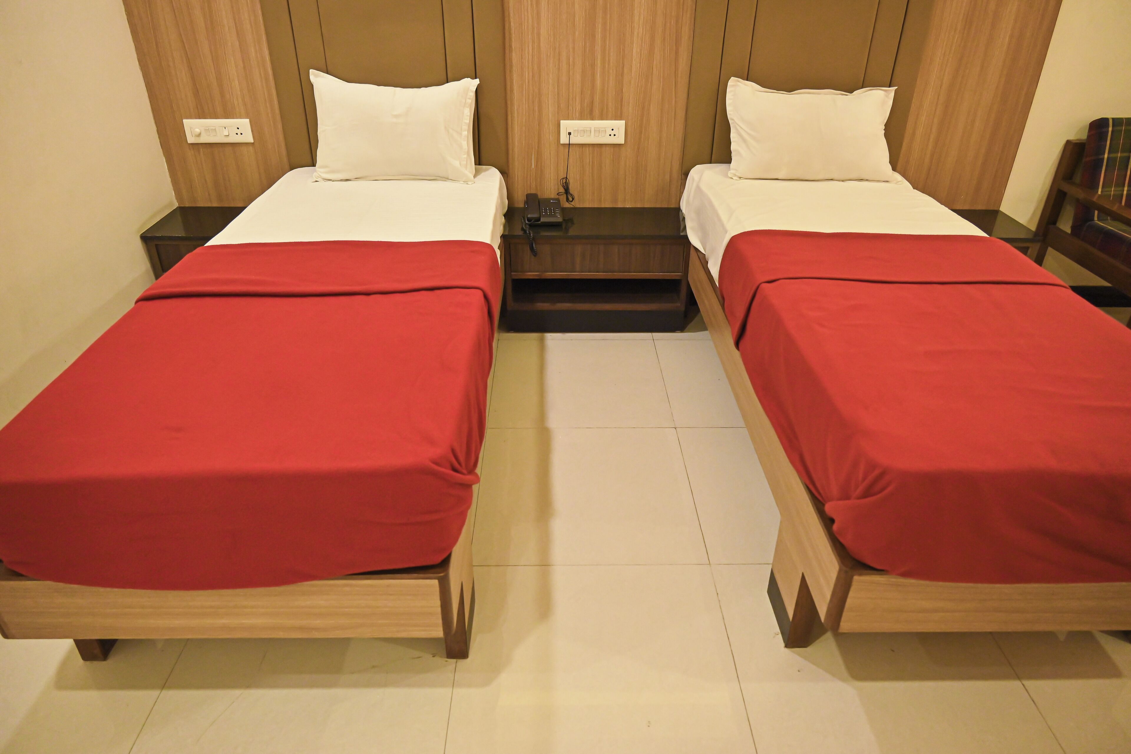 Quarto duplo com duas camas de solteiro, cada uma com colcha vermelha e almofadas brancas, uma pequena mesa de cabeceira com telefone e uma tomada na parede.