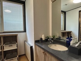 Casa de banho com um espelho grande, uma pia e uma janela com persianas. Existem cestos no chão e produtos de higiene pessoal no balcão.