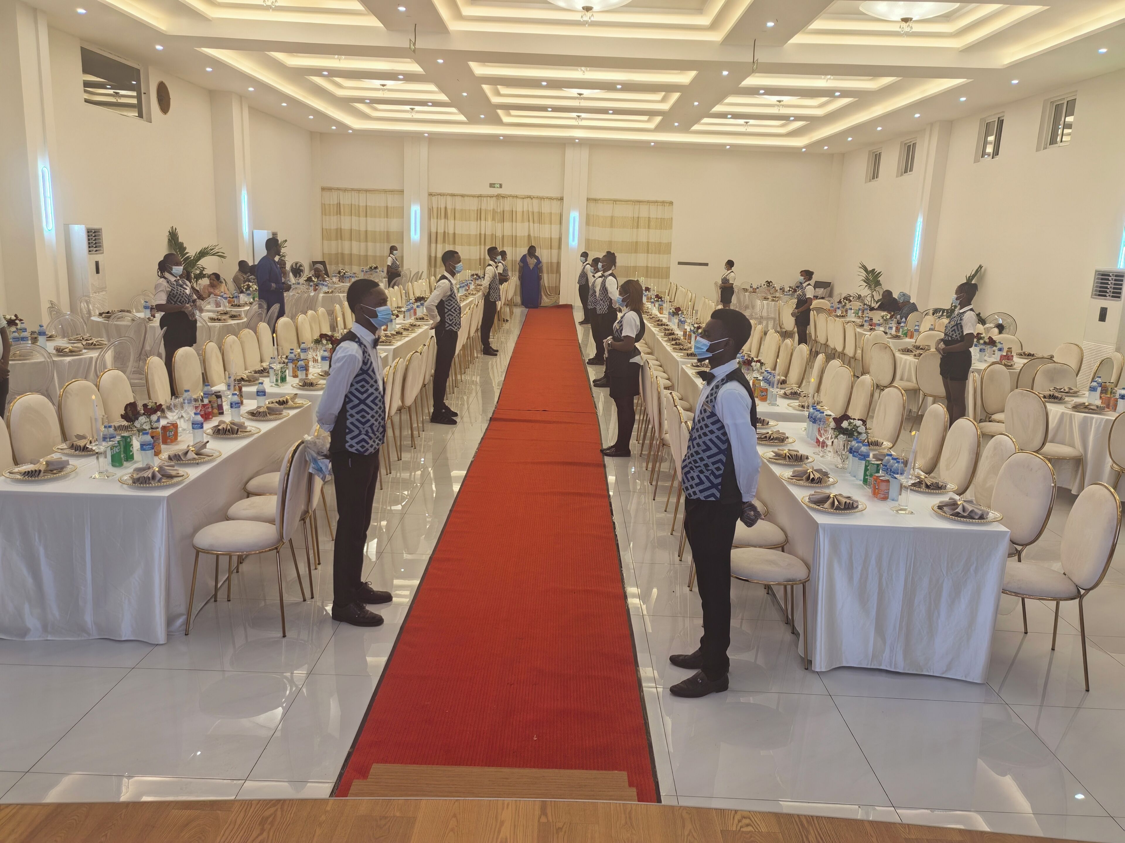 Salão de banquetes com mesas montadas para um evento formal, funcionários uniformizados e um tapete vermelho.