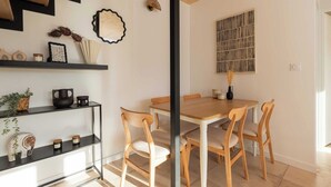 Uma área de refeições com uma mesa e cadeiras de madeira, uma estante de metal preta com objetos decorativos e uma luminária fixa na parede.