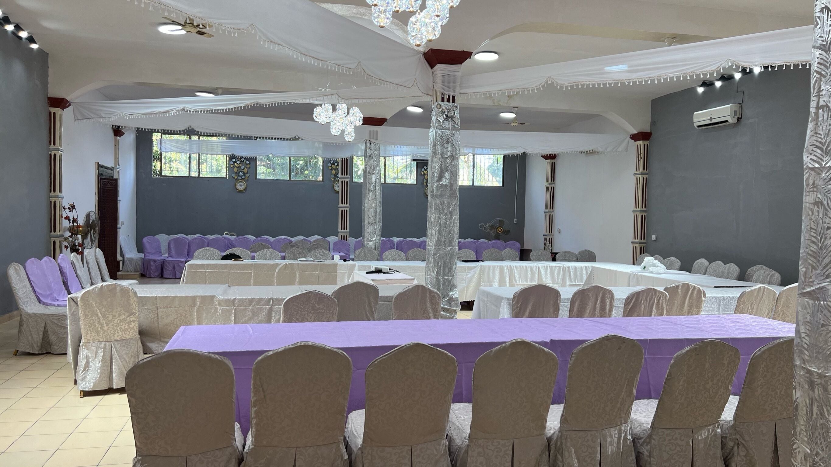 Uma sala de banquetes com várias mesas redondas cobertas por toalhas brancas e capas de cadeiras roxas.