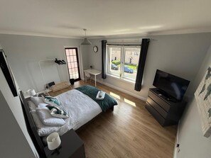 Quarto com cama, mesa de cabeceira, luminária, televisão e janela com vista para a rua.