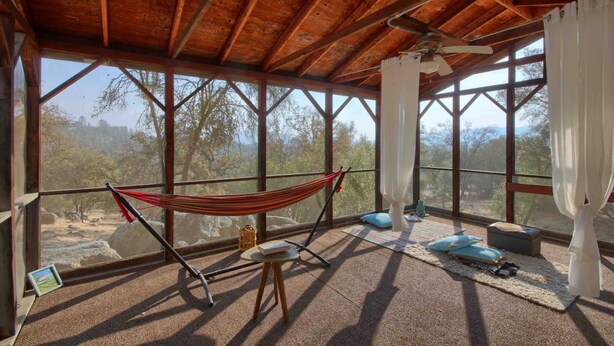 Um quarto espaçoso com teto de madeira, grandes janelas, uma rede, um ventilador de teto e vista para árvores e pedras lá fora.
